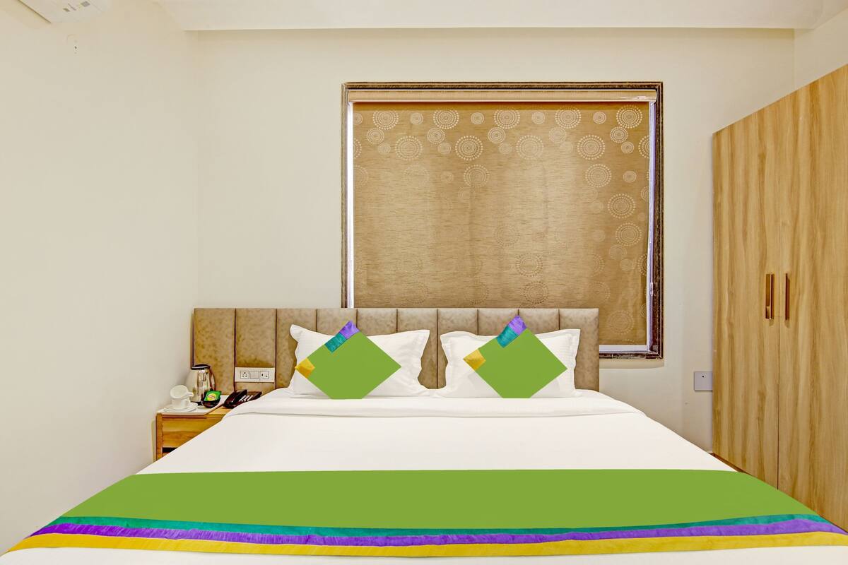 Quarto de hotel com cama, mesa de cabeceira, telefone e janela com cortina.
