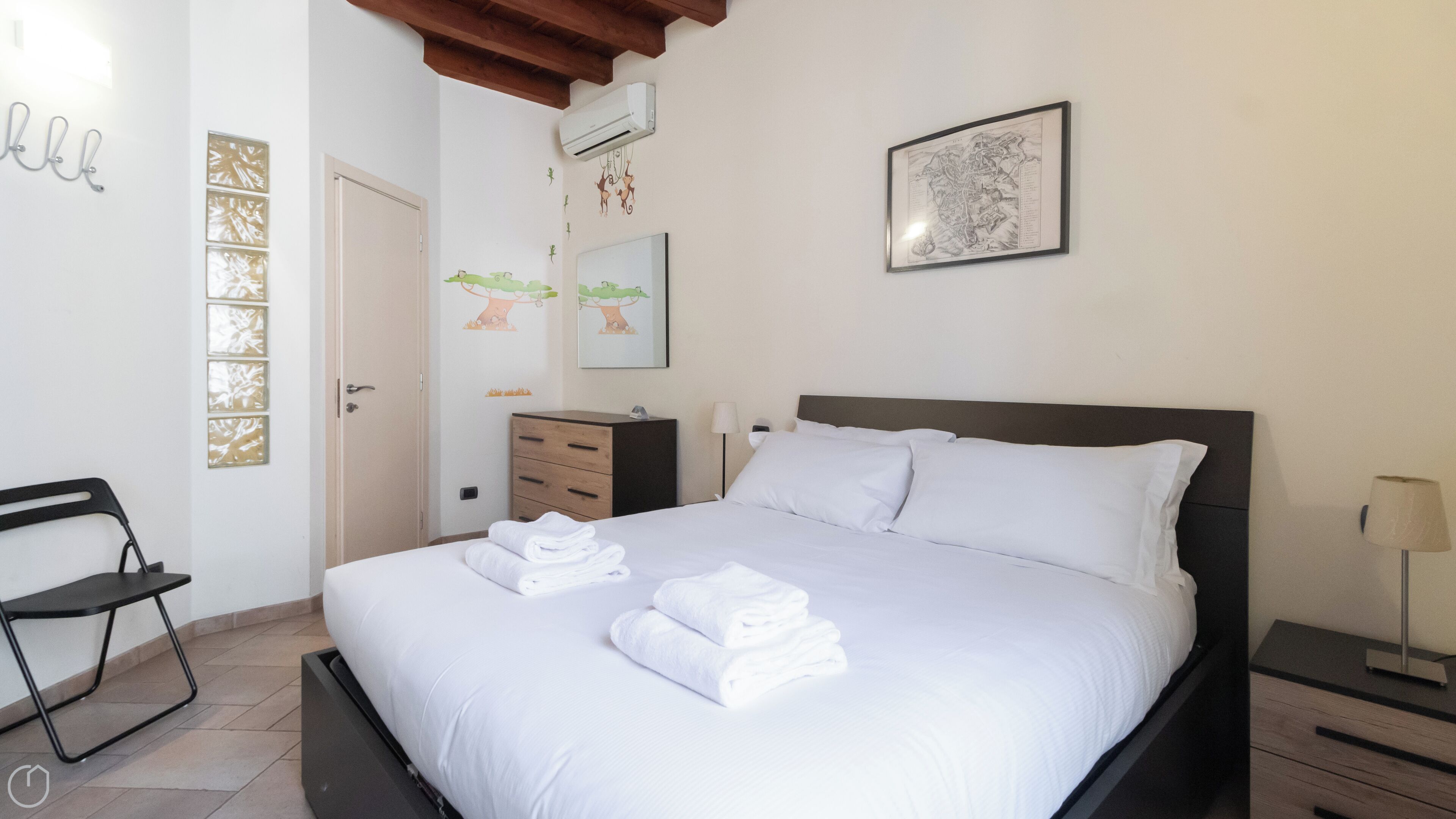 Uma cama bem arrumada com roupa de cama e toalhas brancas, cabeceira de madeira, mesa de cabeceira pequena com abajur e ar-condicionado fixado na parede.