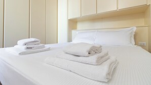 Cama bem arrumada com lençóis e toalhas brancas, cabeceira branca e parede lisa com painéis verticais.