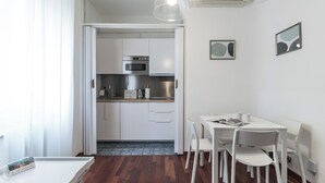 Cozinha compacta com armários brancos, micro-ondas e máquina de café. Há uma mesa de jantar com cadeiras e um aviso de 'Proibido Fumar'.