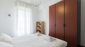 Quarto com uma cama grande, guarda-roupa de madeira, estante de livros e janela com cortinas finas.