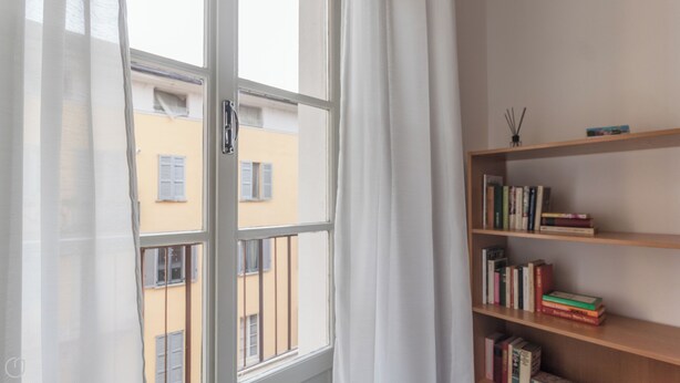 Quarto com uma janela grande, uma estante de livros e vista para um edifício amarelo lá fora.