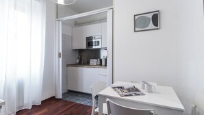 Cozinha compacta com armários brancos, micro-ondas e cafeteira. Há uma mesa de jantar com cadeiras e um quadro na parede.