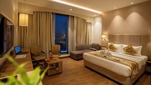 Quarto de hotel moderno com uma cama grande, uma mesa com um laptop, um sofá e uma TV.
