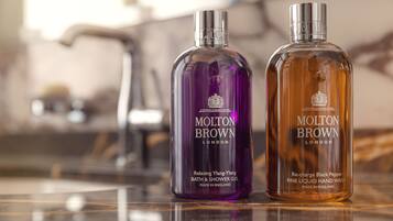 Duas embalagens de gel de banho e duche Molton Brown sobre uma superfície refletora.