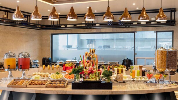 Disposição de buffet com diversos alimentos e bebidas, cozinha moderna ao fundo e iluminação pendente.