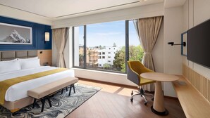 Quarto de hotel com uma cama grande, uma escrivaninha com cadeira, uma televisão e uma janela com vista para a cidade.
