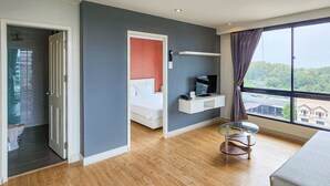 Quarto de hotel moderno com piso de madeira, cama com roupa de cama branca, televisão em prateleira branca e uma janela grande com vista para edifícios e árvores.