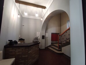 Uma área de recepção com balcão de tijolos, escada com corrimão vermelho e corredor que leva a uma porta.