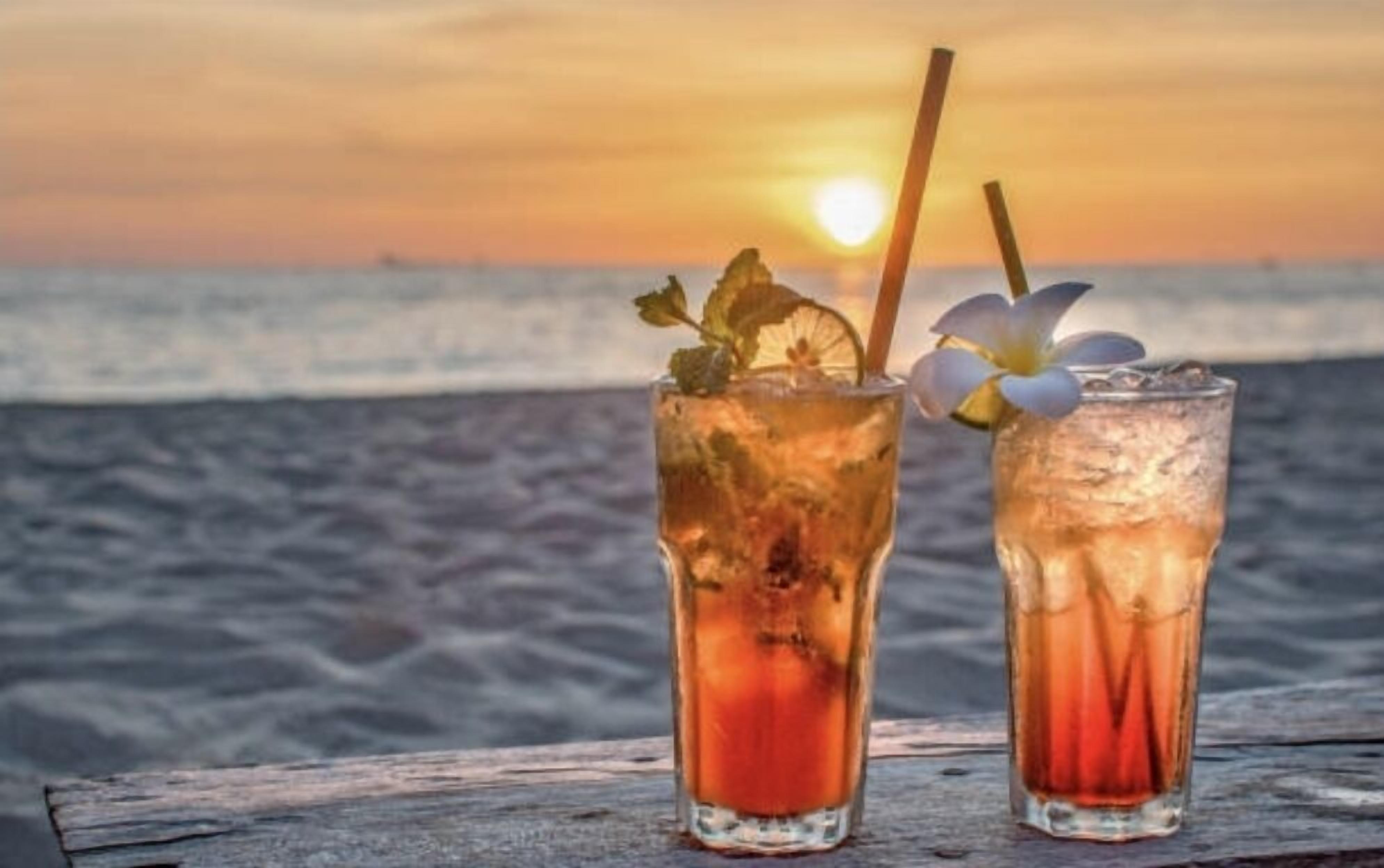 Duas bebidas geladas com canudos sobre uma superfície de madeira à beira-mar ao pôr do sol.