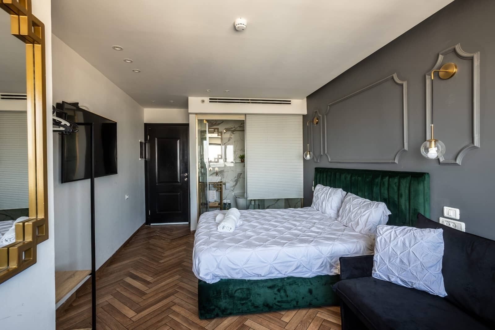 Quarto de hotel moderno com cama grande, um banco estofado verde, televisão de tela plana e um espelho.