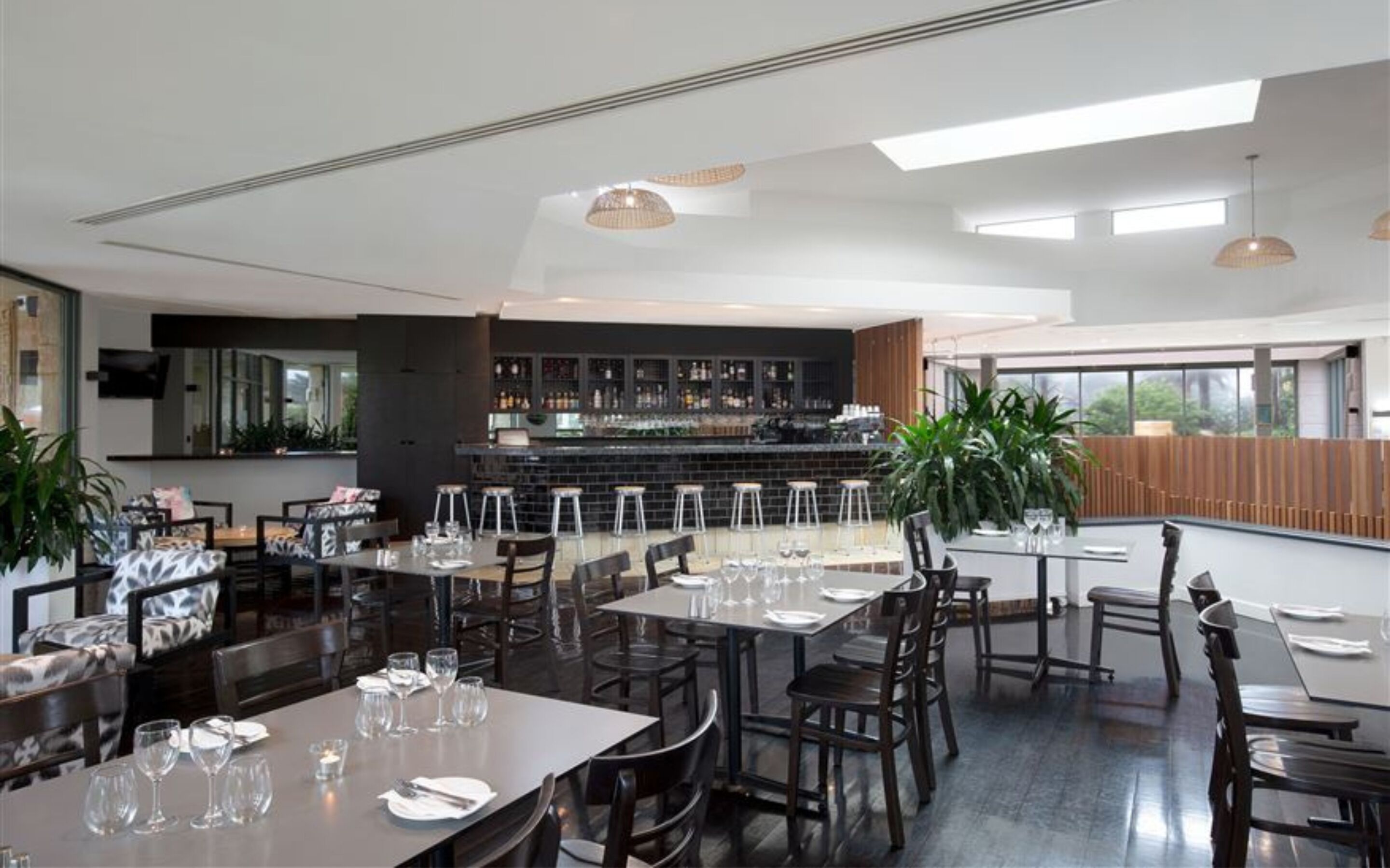 Restaurante moderno com pé-direito alto, área de bar e grandes janelas que proporcionam luz natural.