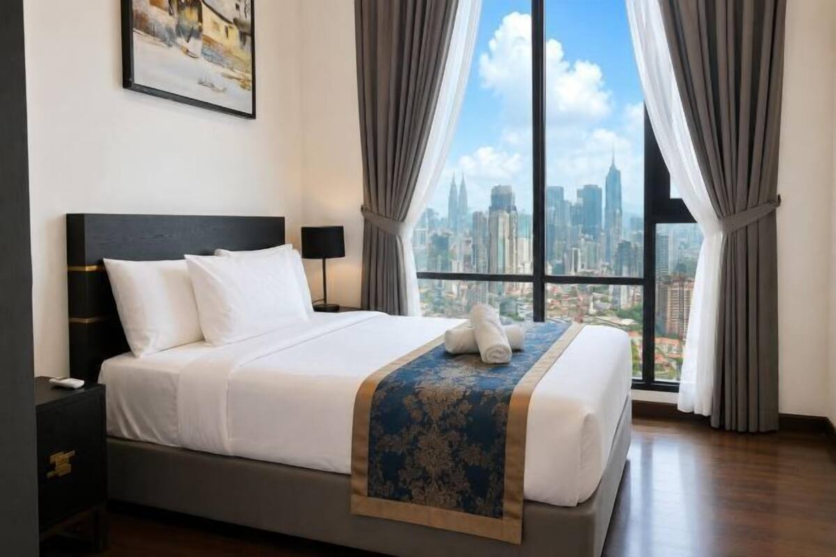 Quarto de hotel com cama, mesa de cabeceira, luminária e uma janela grande com vista para a cidade.