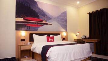 Quarto de hotel com uma cama grande, mesa de cabeceira, escrivaninha e televisão fixada na parede.