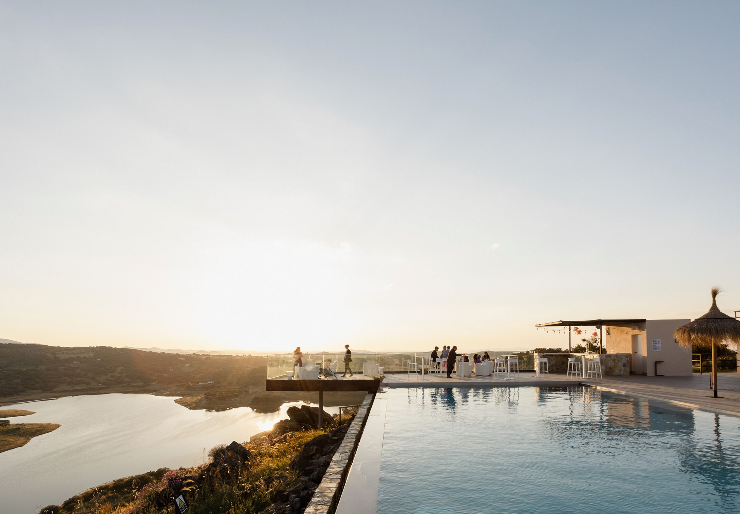 Piscina com vista para um lago, área de estar ao ar livre e um edifício com telhado de palha.