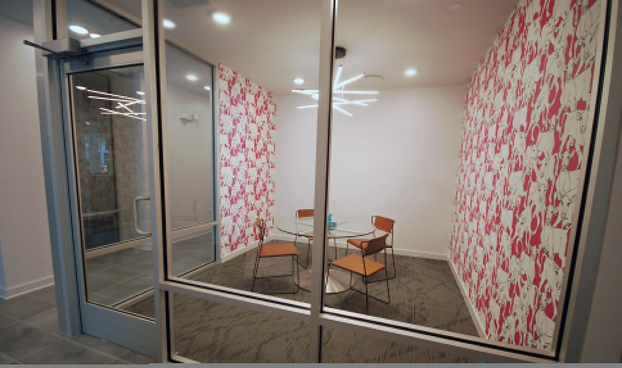 Uma sala de reunião moderna com paredes de vidro, duas cadeiras, uma mesa e um candeeiro no teto.