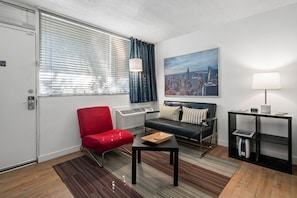 Quarto de hotel moderno com uma cadeira vermelha, um sofá preto, uma pintura de paisagem urbana, um abajur e uma mesa pequena.