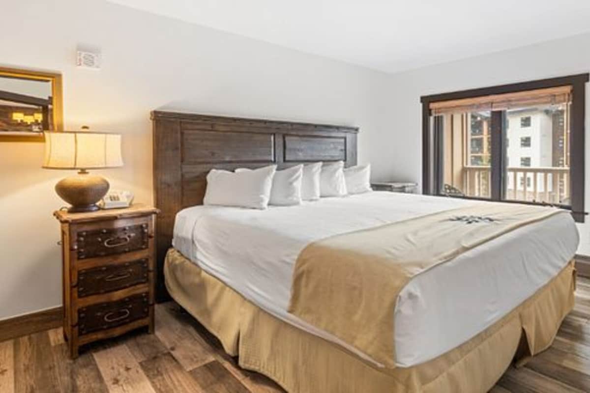 Um quarto com uma cama de madeira, uma mesa de cabeceira com abajur, uma janela com vista e uma TV fixada na parede.