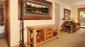 Sala de estar com mesa consola de madeira, um quadro de paisagem emoldurado, luminária e sofá com almofadas.