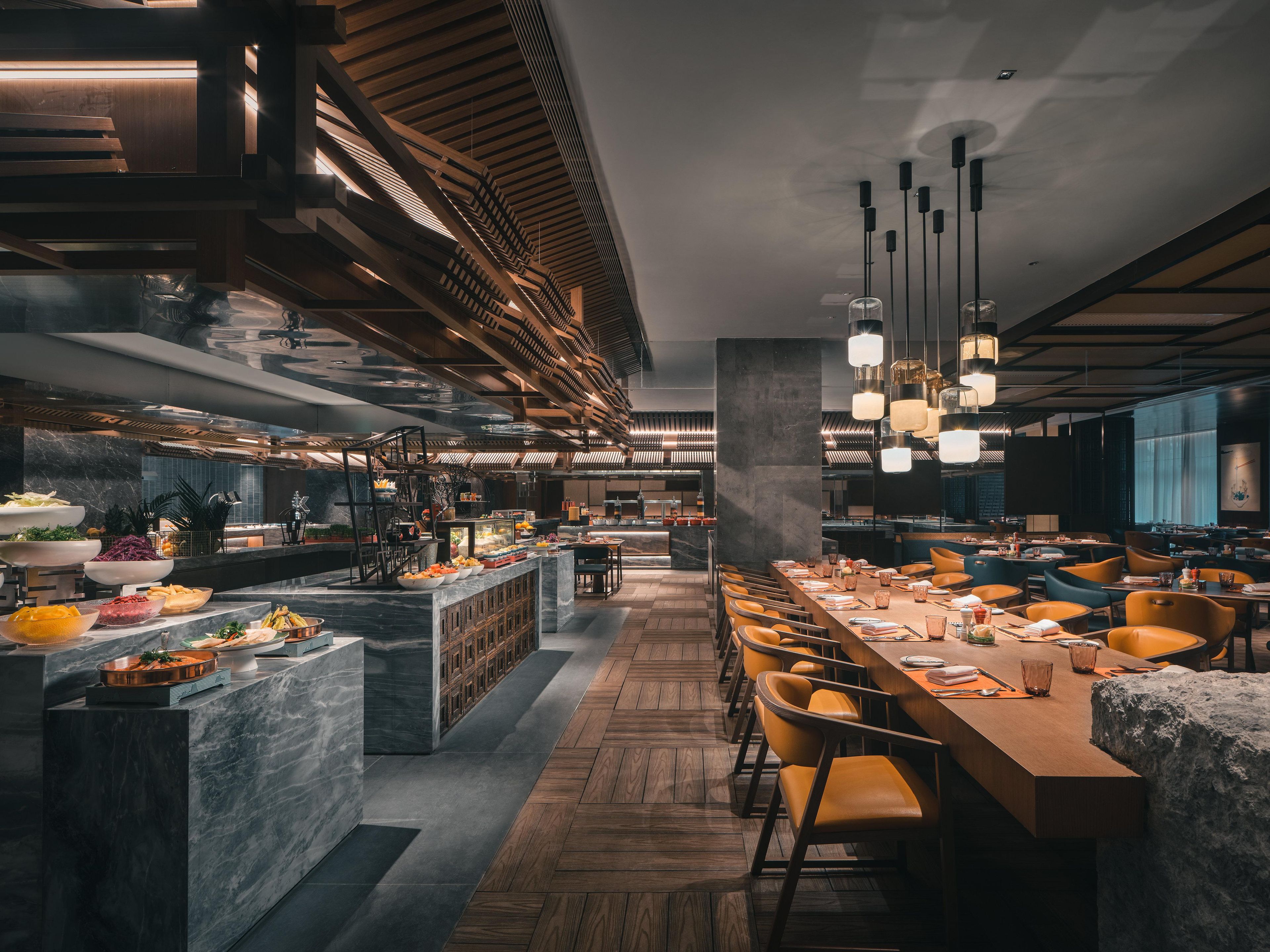 Interior de restaurante moderno com uma longa mesa comunitária, diversas áreas de estar e um balcão central de bar com displays de comida.