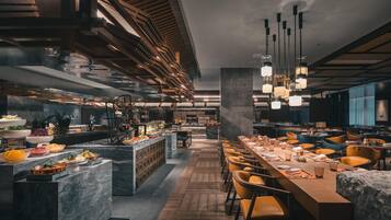 Interior de restaurante moderno com uma longa mesa comunitária, diversas áreas de estar e um balcão central de bar com displays de comida.