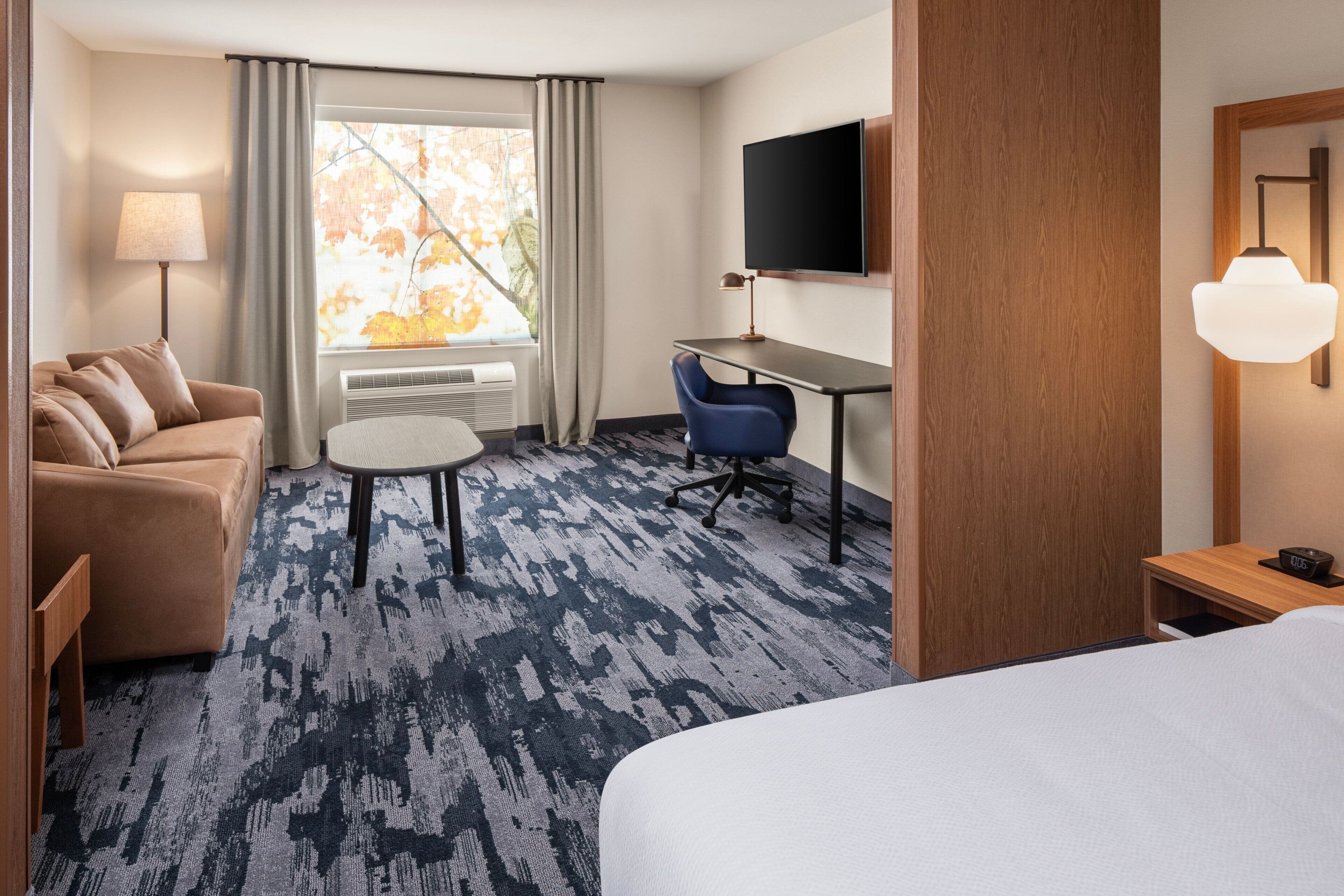 Quarto de hotel com uma cama, uma escrivaninha, uma cadeira, uma televisão e um abajur.