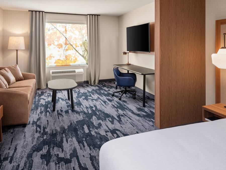 Quarto de hotel com uma cama, uma escrivaninha, uma cadeira, uma televisão e um abajur.
