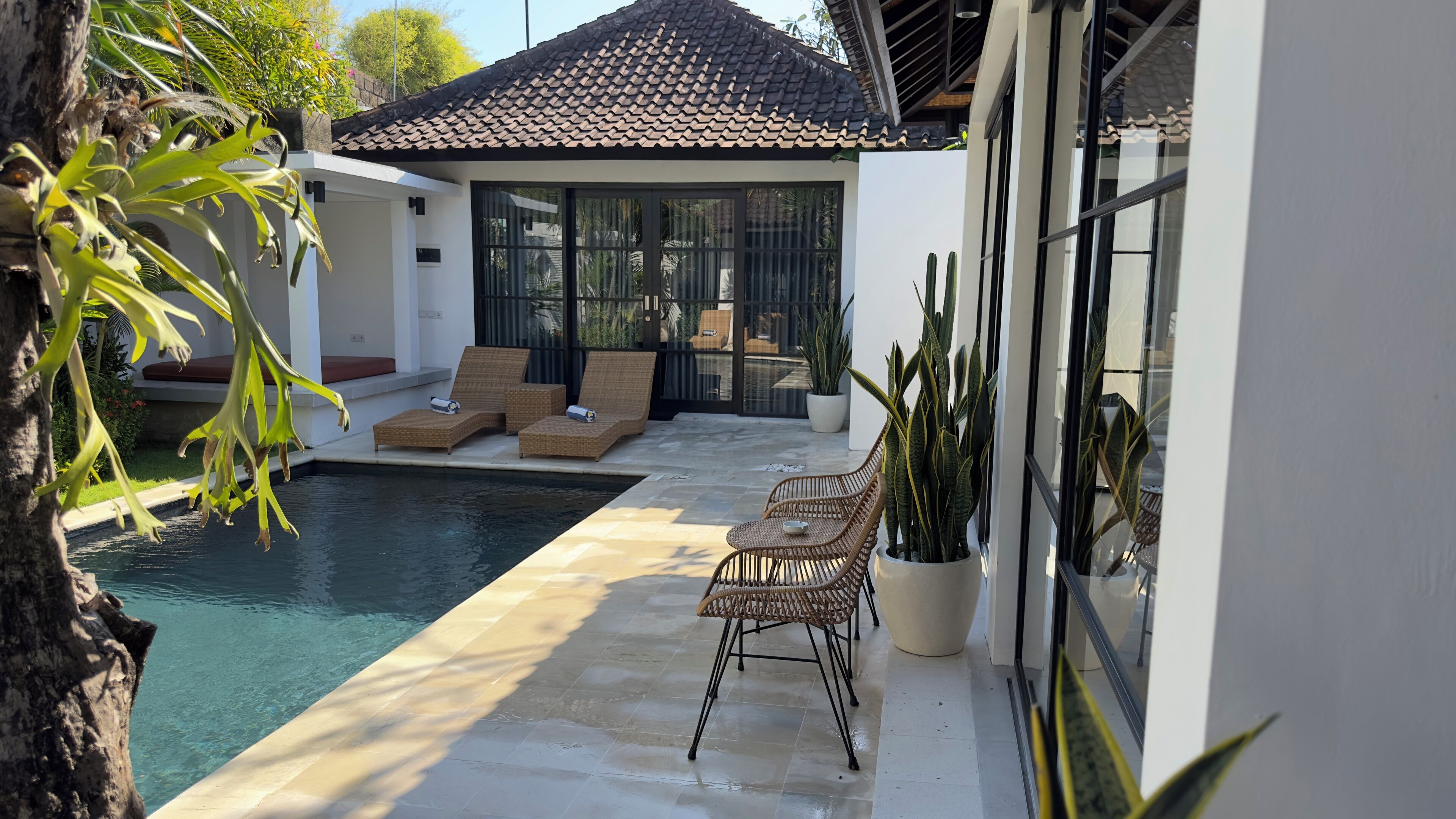 Uma área de piscina moderna ao ar livre, com espreguiçadeiras, um vaso de planta e um terraço envidraçado.