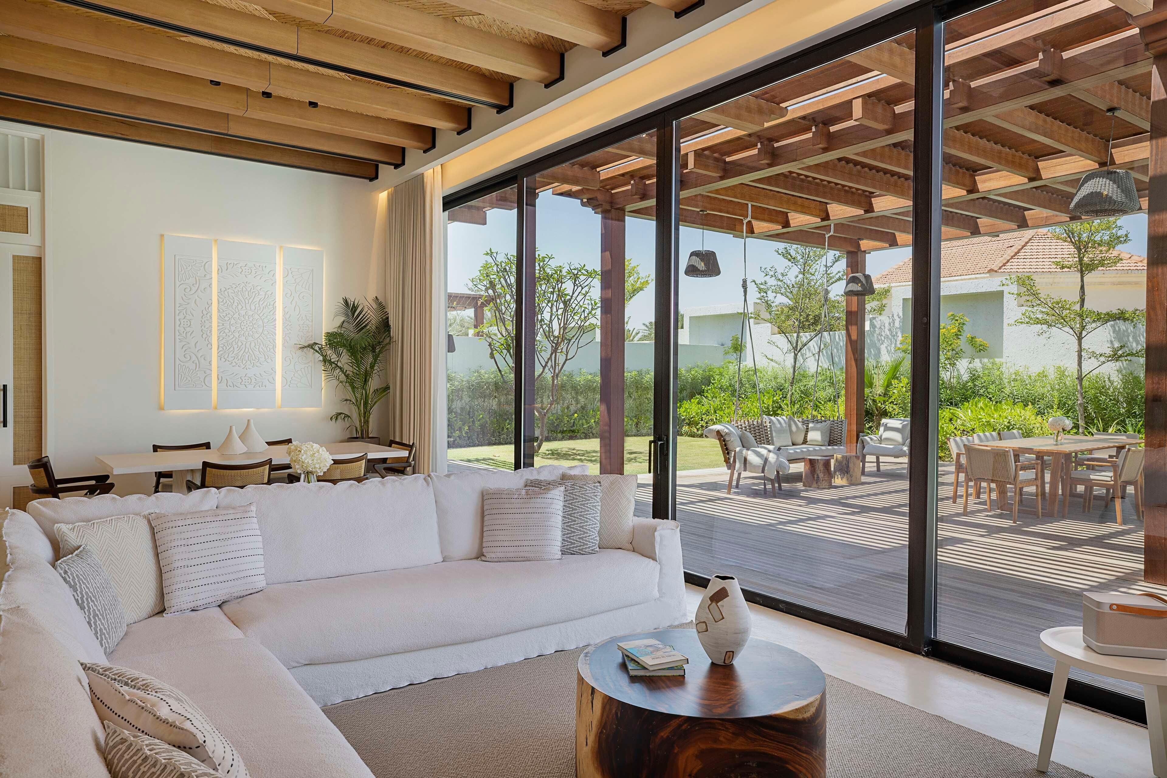 Uma sala de estar moderna com um sofá branco grande, uma mesa de centro redonda de madeira e vista para um deck externo com móveis.