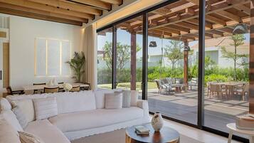 Uma sala de estar moderna com um sofá branco grande, uma mesa de centro redonda de madeira e vista para um deck externo com móveis.
