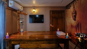 Um quarto com uma mesa de massagem de madeira, uma TV montada, uma pintura de Buda e velas.