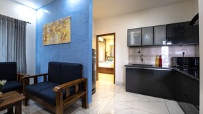 Um espaço de estar compacto com uma parede azul de destaque, um sofá de madeira, uma pequena mesa de jantar, uma cozinha com armários pretos e um micro-ondas, e uma pintura na parede.