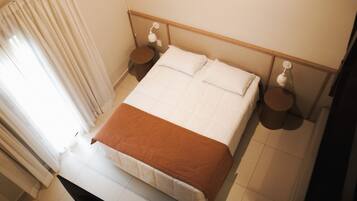 Cama bem arrumada com roupa de cama branca e castanha, duas mesas de cabeceira com abajures e uma janela com cortinas.