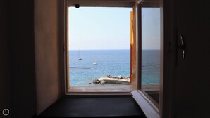 Vista através de uma porta para uma varanda com vista para um mar calmo, com barcos e um cais rochoso.
