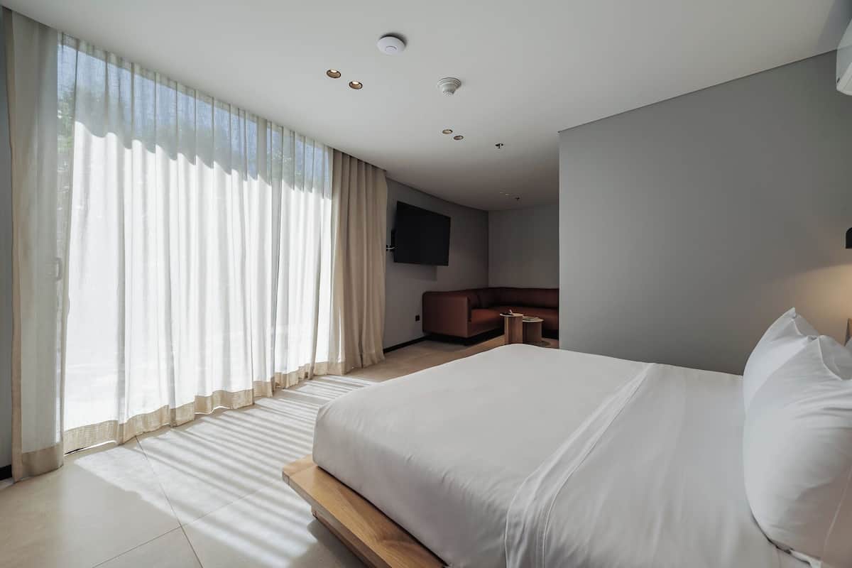 Quarto de hotel moderno com uma cama grande, área de estar com sofá e uma mesa pequena, e uma TV fixada na parede.