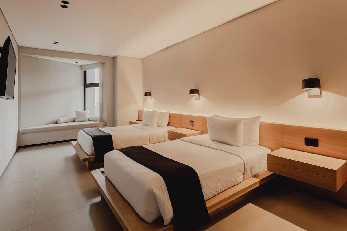 Quarto de hotel moderno com duas camas, um banco embutido, mesas de cabeceira e iluminação na parede.