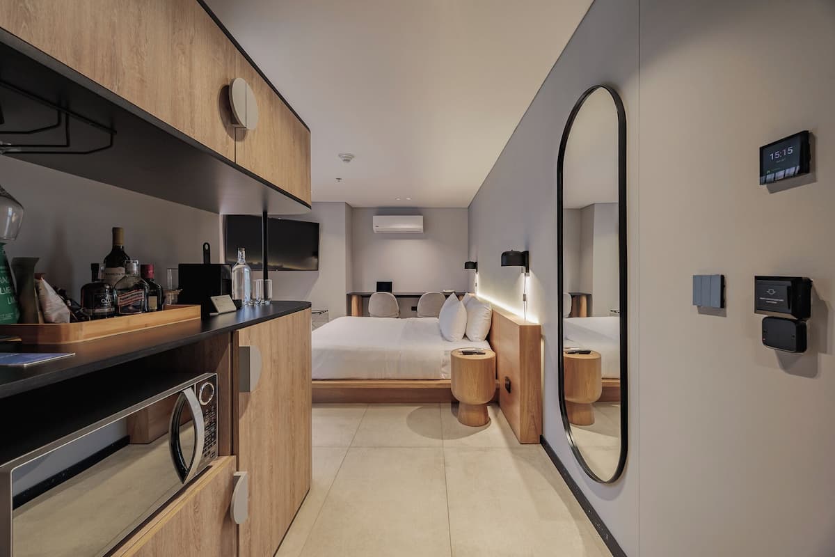 Quarto de hotel moderno com escrivaninha de madeira, minibar e uma cama grande com cabeceira.