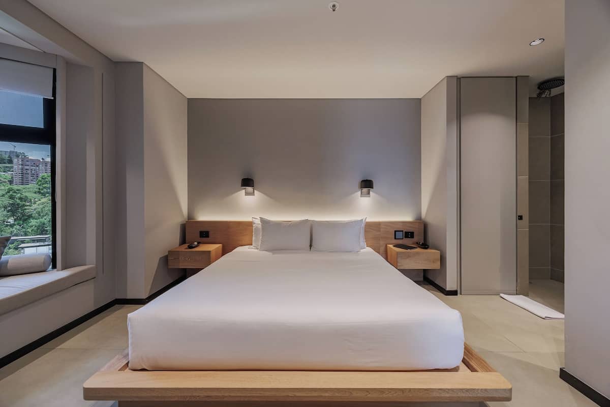 Quarto de hotel moderno com uma cama grande, duas mesas de cabeceira, uma luminária de cabeceira, uma janela com persianas e vista para edifícios e árvores.