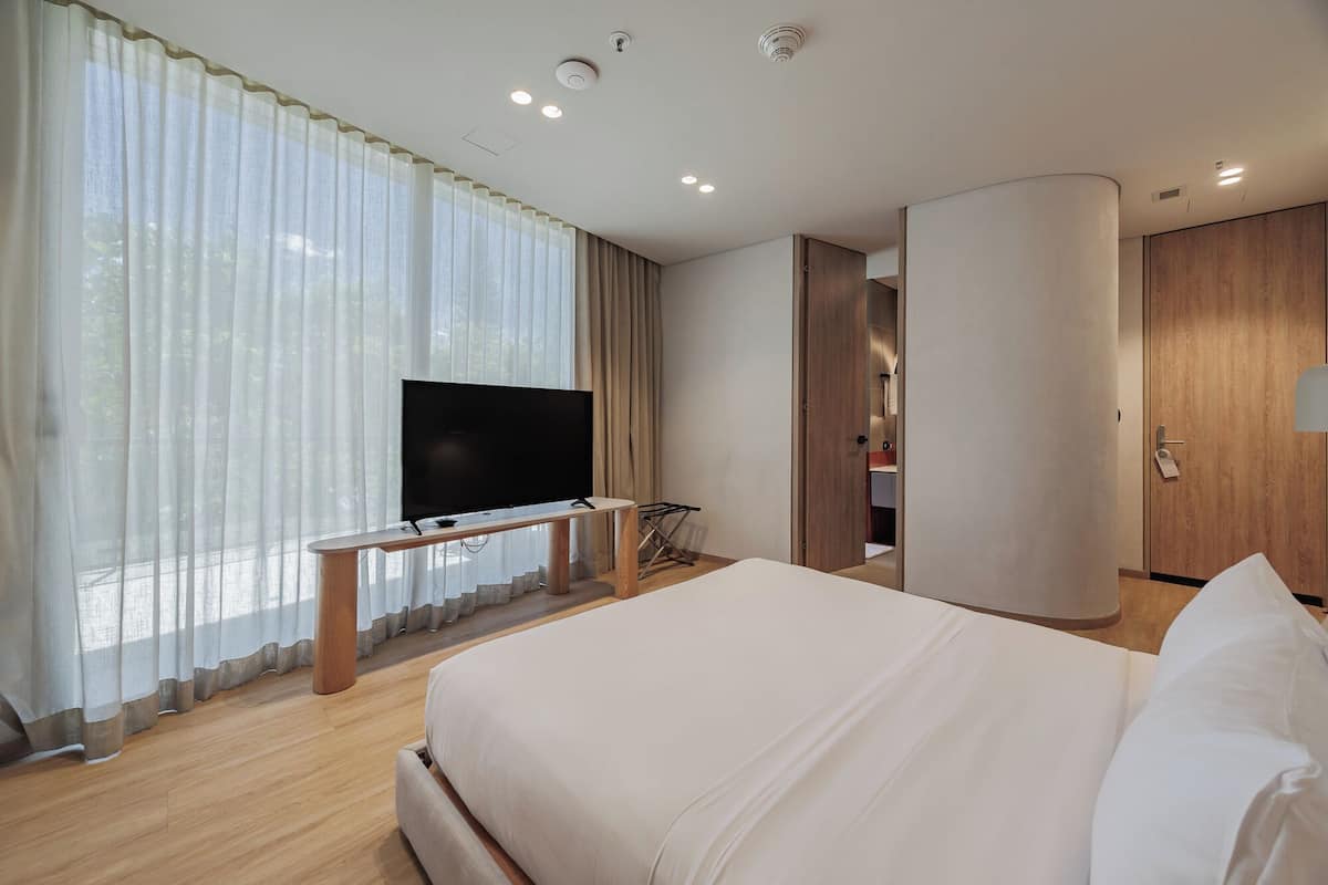 Quarto de hotel moderno com uma cama grande, uma televisão de tela plana em um suporte de madeira, um guarda-roupa e um banheiro visível por uma porta aberta.