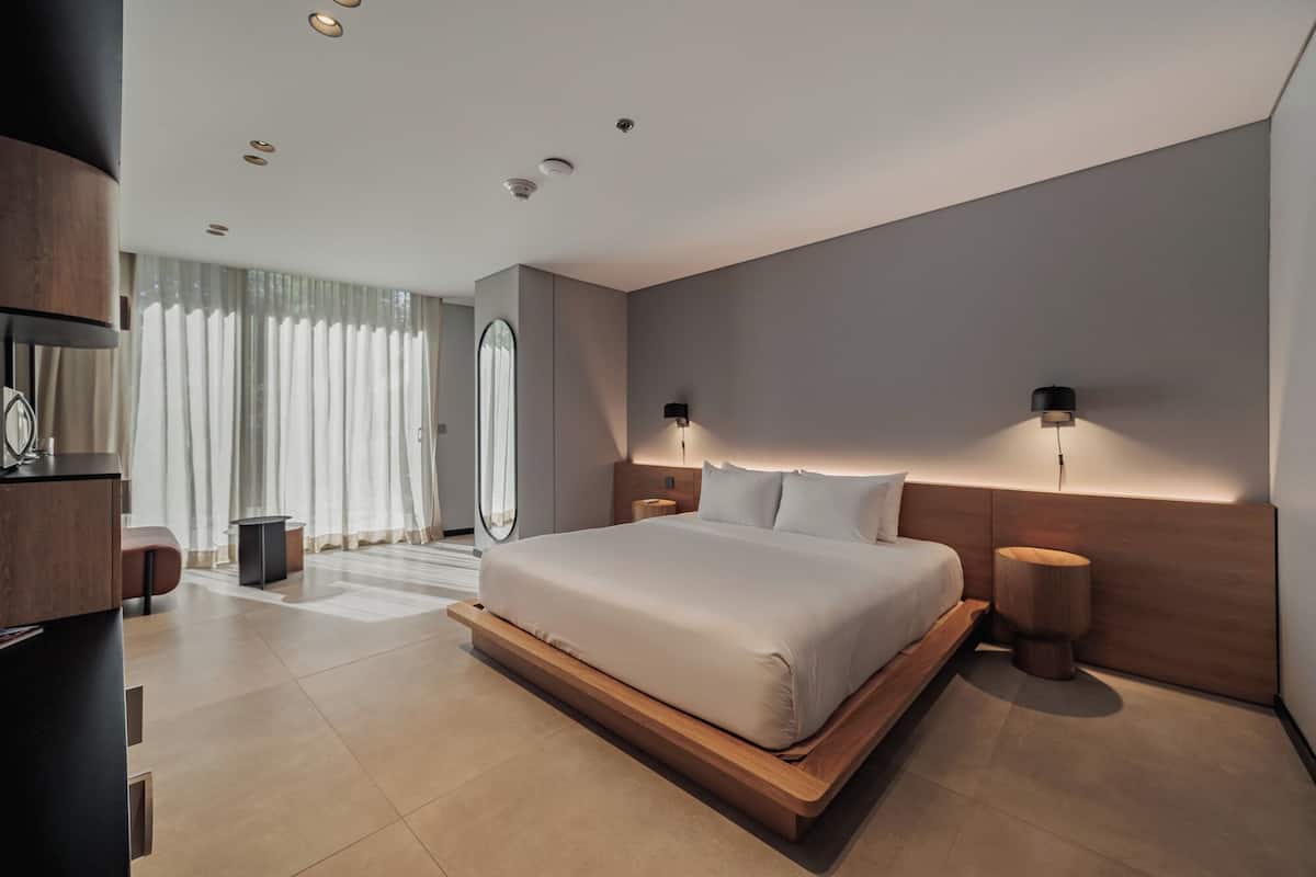 Quarto de hotel moderno com uma cama grande, duas mesas de cabeceira, um espelho e vista para o exterior através de cortinas finas.