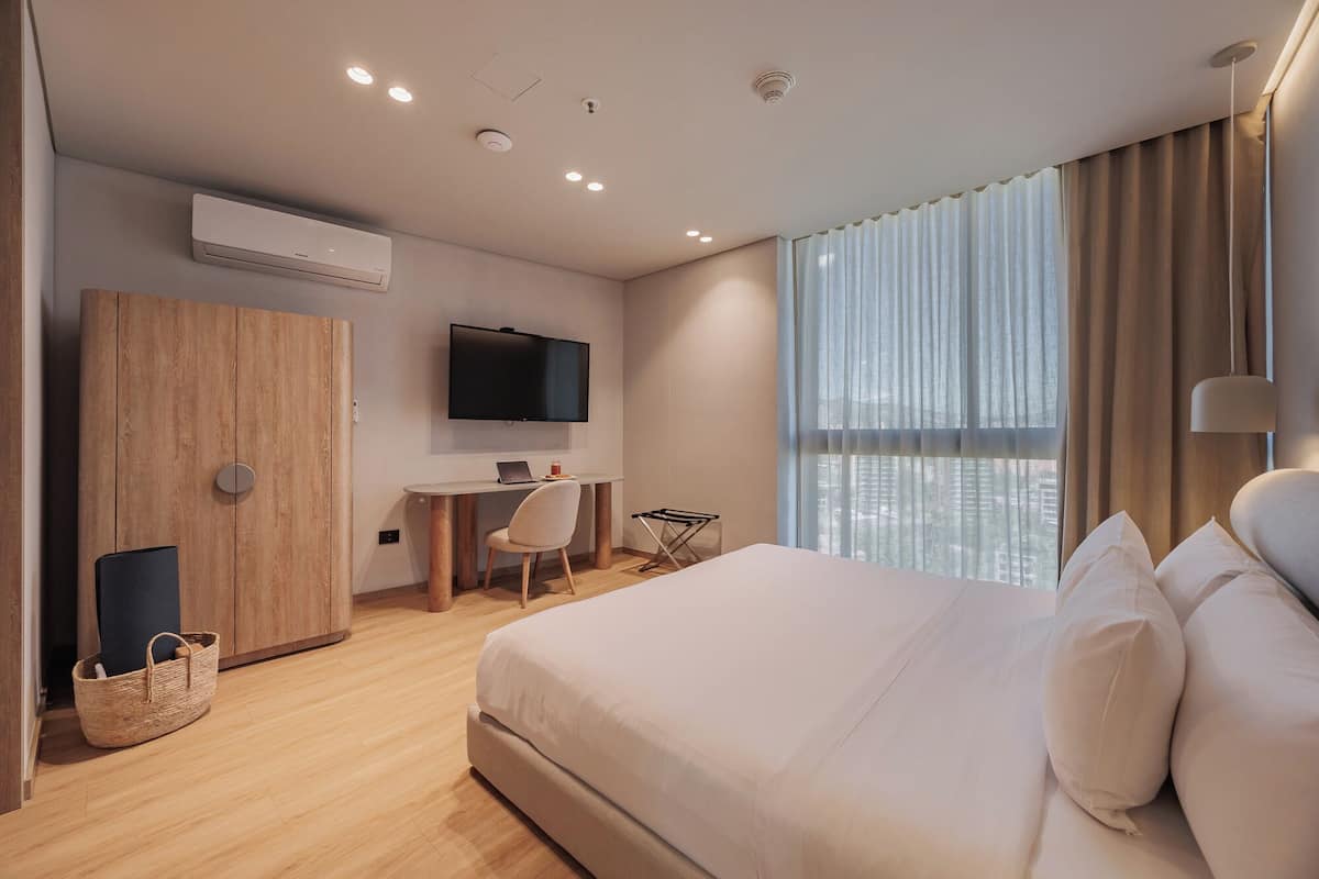Quarto de hotel moderno com uma cama grande, uma mesa com um laptop, uma televisão e vista da cidade através de amplas janelas.