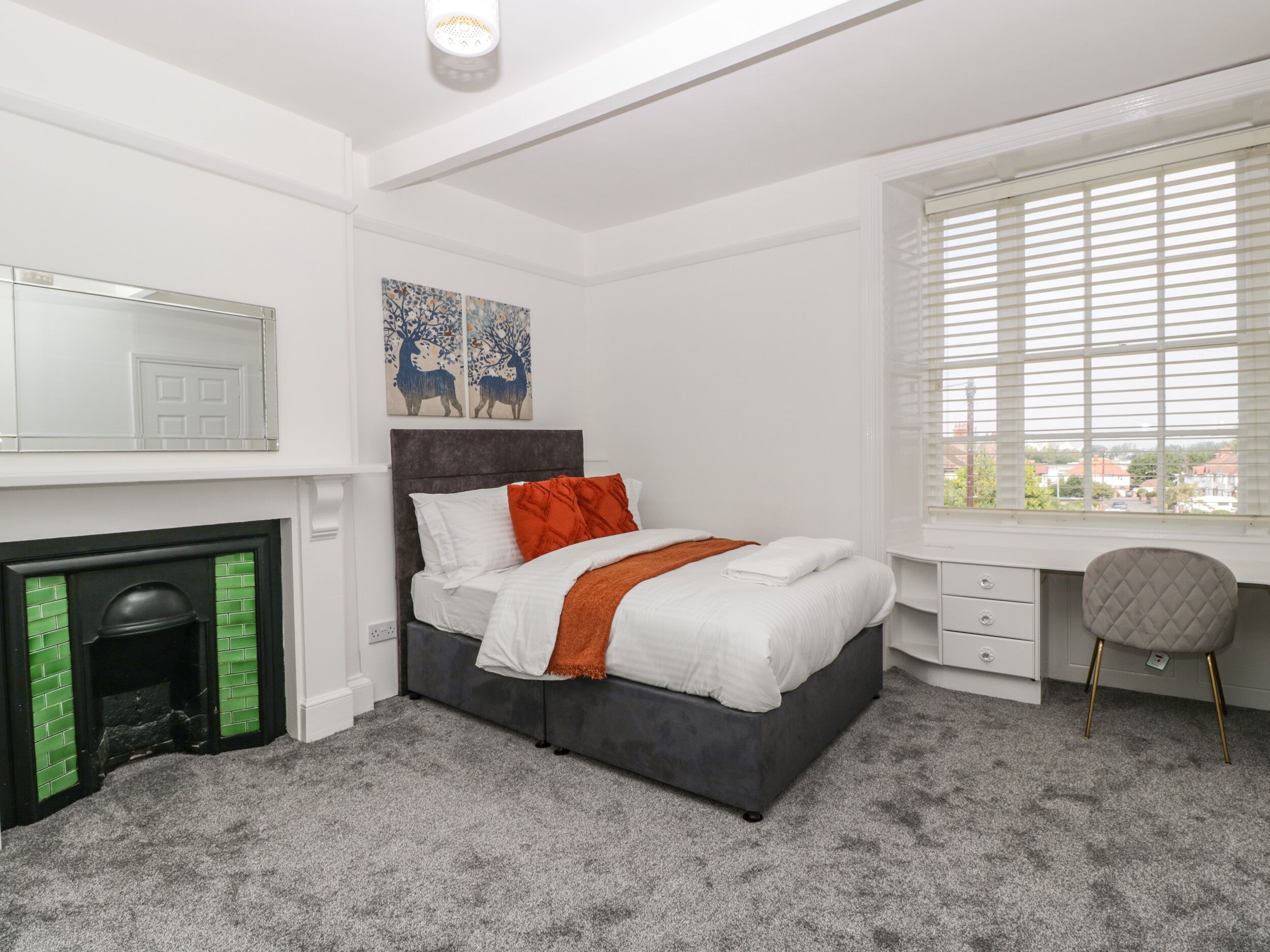 Um quarto com cama, escrivaninha, poltrona, lareira e janela com persianas.