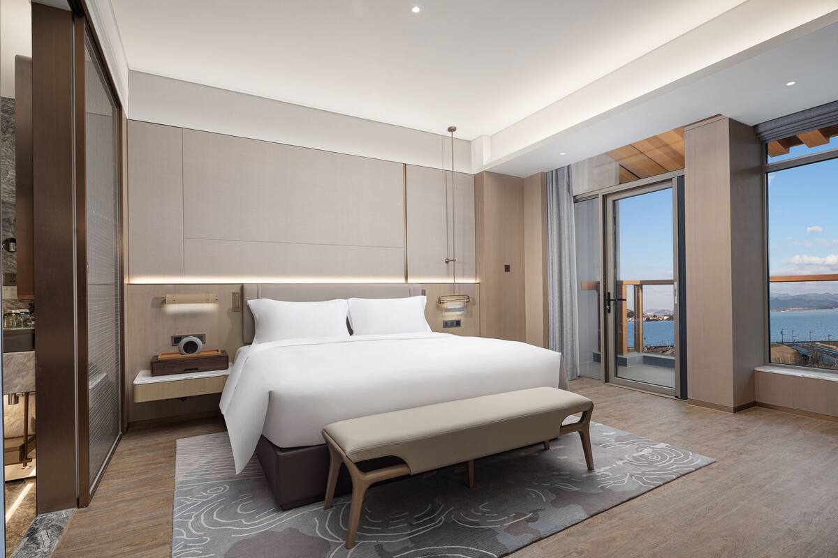 Quarto moderno com uma cama grande, um banco, uma mesa de cabeceira e vista para o mar através de uma porta de correr.