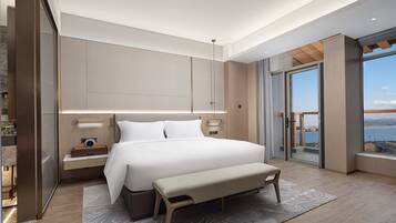 Quarto moderno com uma cama grande, um banco, uma mesa de cabeceira e vista para o mar através de uma porta de correr.