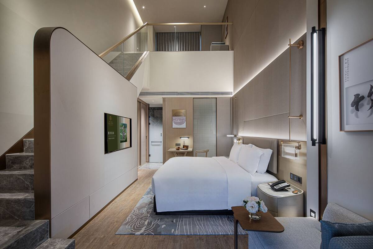 Quarto de hotel moderno com uma cama grande, uma escrivaninha, uma mesinha com um vaso e uma escada que leva a um andar superior.