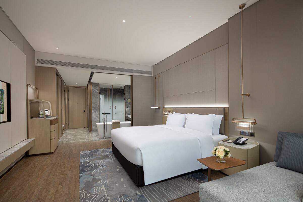 Quarto de hotel moderno com uma cama grande, mesas de cabeceira, uma escrivaninha e um banheiro visível por uma porta aberta.