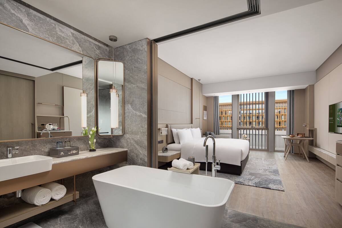 Quarto de hotel moderno com uma banheira grande, toucador com espelho, cama com vista para a cidade e uma pequena mesa com abajur.