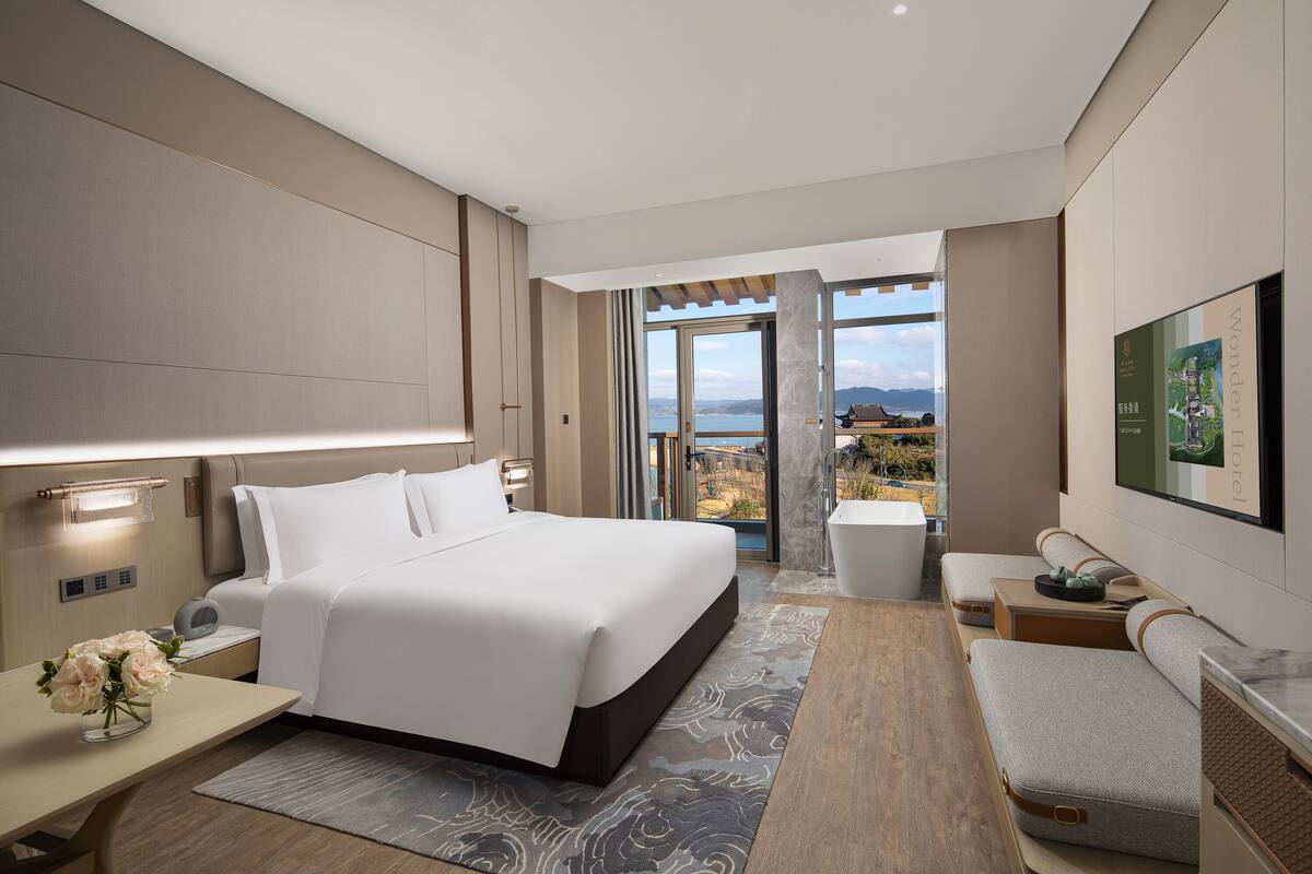 Quarto de hotel moderno com uma cama grande, área de estar, televisão e vista para o exterior.