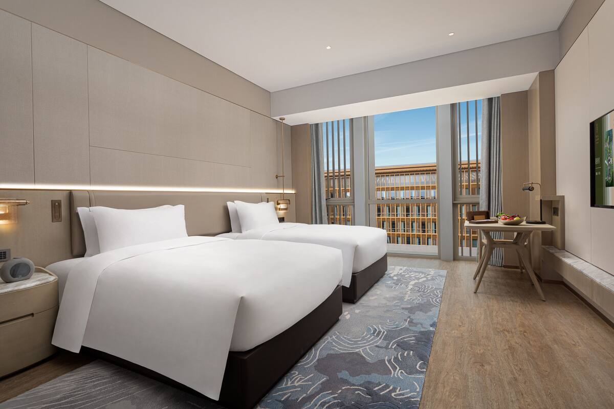 Quarto de hotel moderno com duas camas, uma escrivaninha e uma janela grande com vista para a cidade.