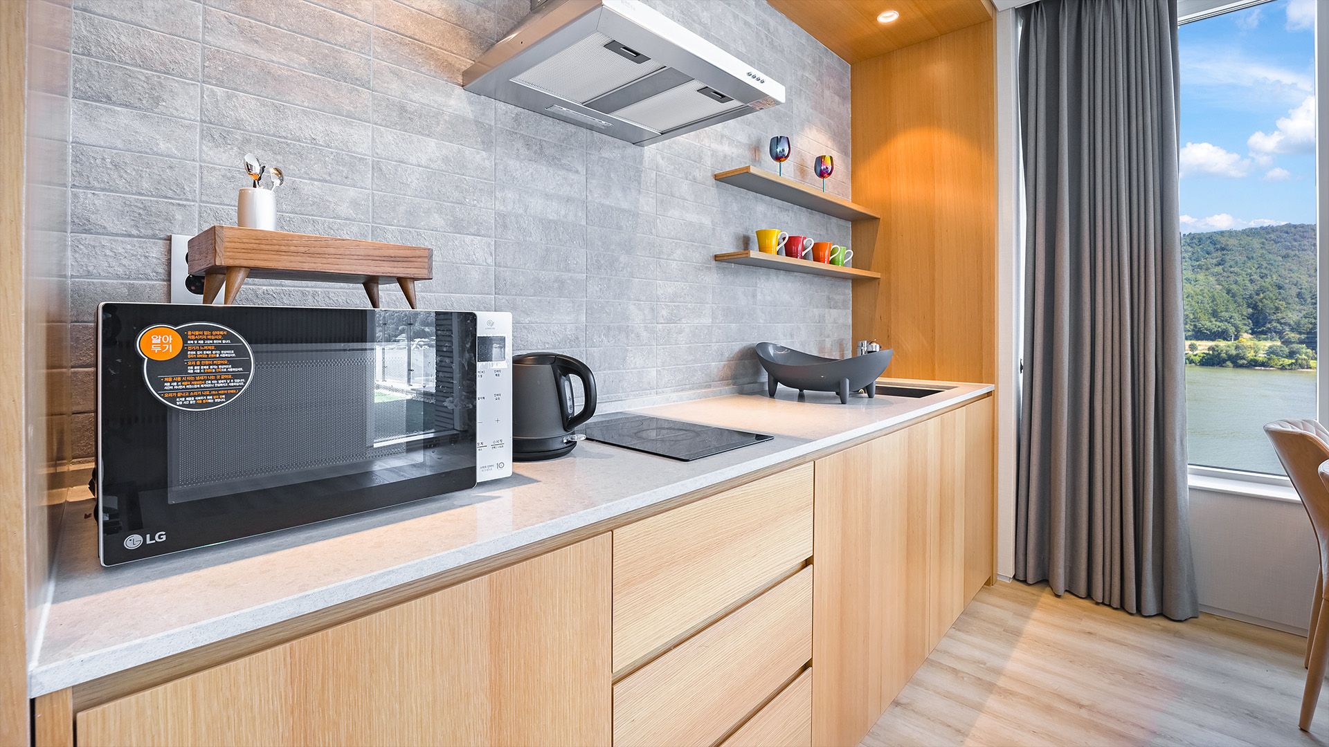 Cozinha moderna com micro-ondas embutido, chaleira e vista para um lago pela janela.